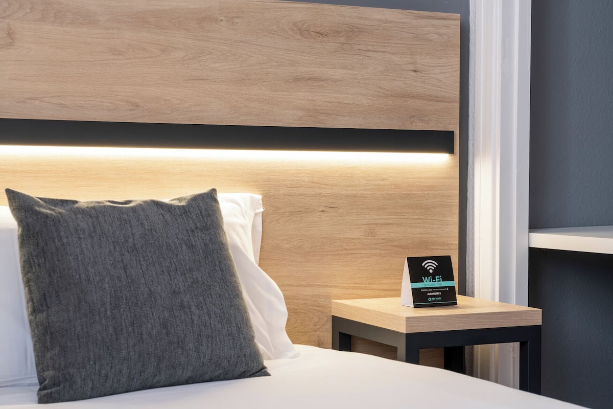 Quarto de hotel com cabeceira de madeira, mesa de cabeceira com sinal de Wi-Fi e travesseiro cinza.