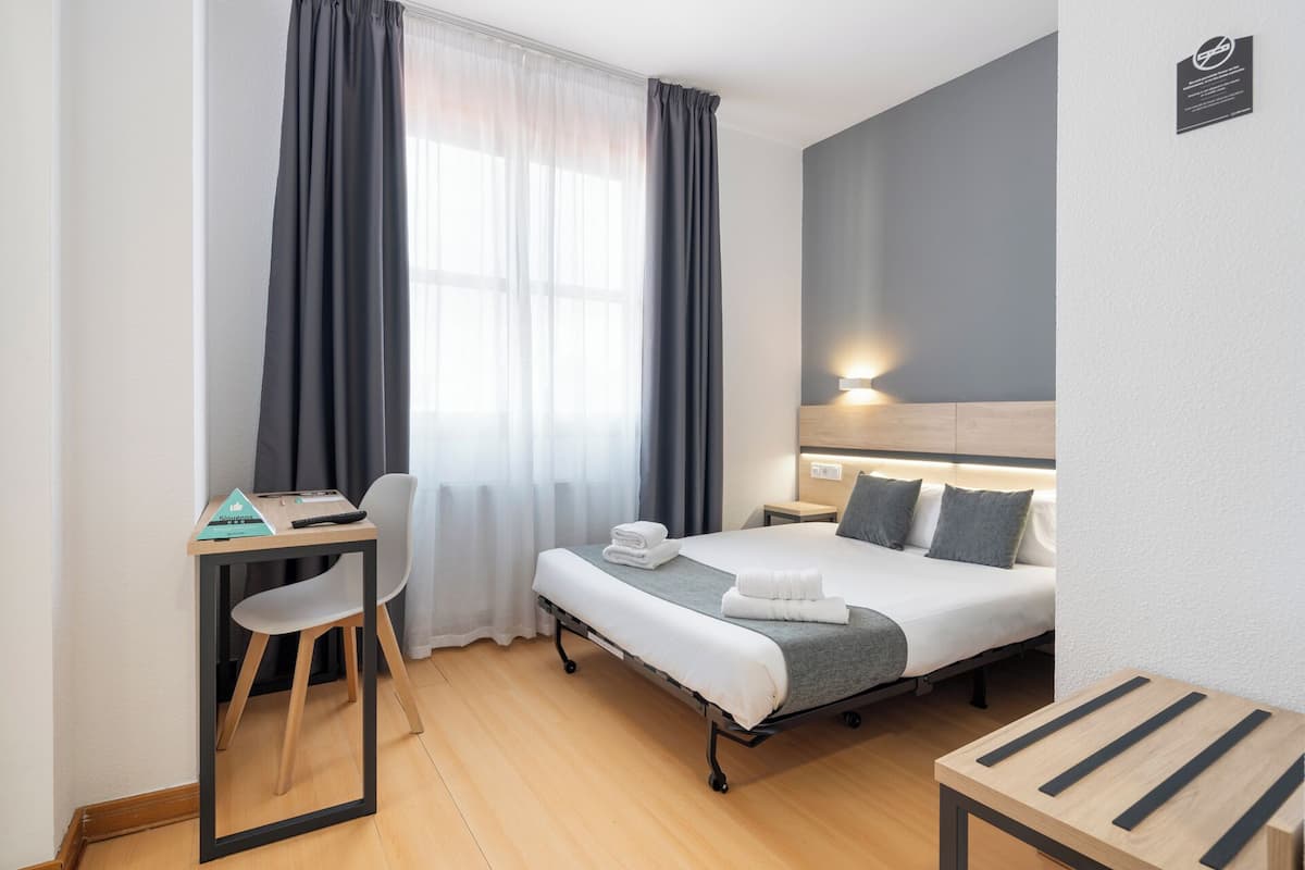 Quarto de hotel com cama, mesa de cabeceira, cadeira, escrivaninha e janela com cortinas.