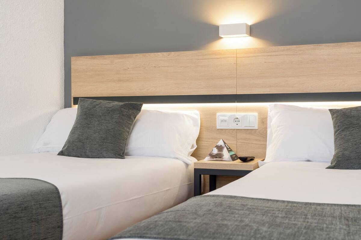 Quarto de hotel com duas camas, uma mesa de cabeceira e um candeeiro fixado na parede.