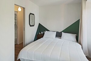 Cama bem arrumada com lençóis brancos, mesa de cabeceira com abajur e parede verde decorativa.
