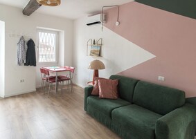 Quarto com sofá verde, mesa de jantar com cadeiras, janela com vista e parede com design geométrico.