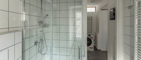 Um banheiro moderno com chuveiro em box de vidro, paredes com azulejos brancos e uma área de serviço visível com máquina de lavar.