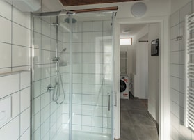 Um banheiro moderno com chuveiro em box de vidro, paredes com azulejos brancos e uma área de serviço visível com máquina de lavar.