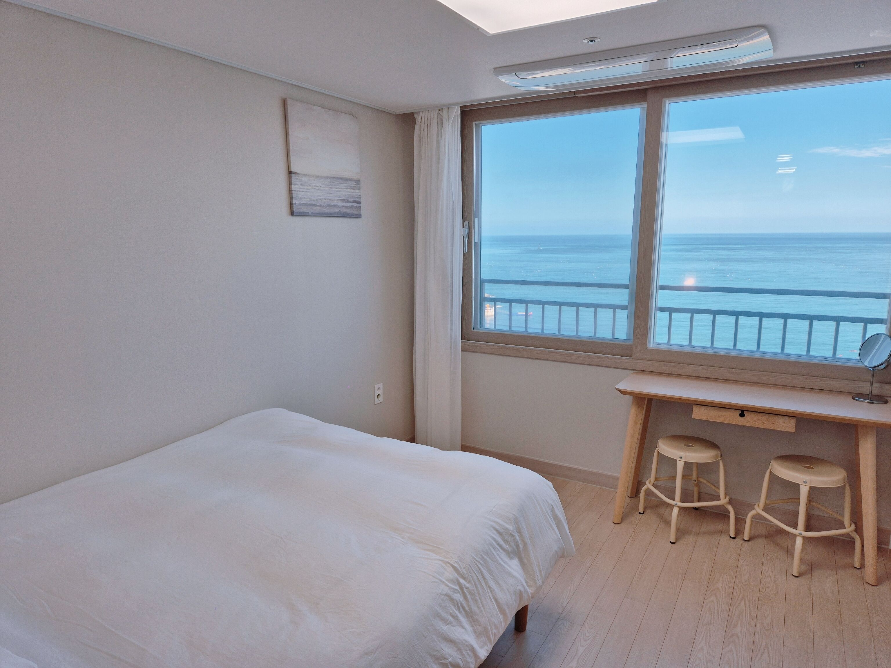 Quarto com uma janela grande, uma cama, uma escrivaninha com dois banquinhos e um quadro na parede.
