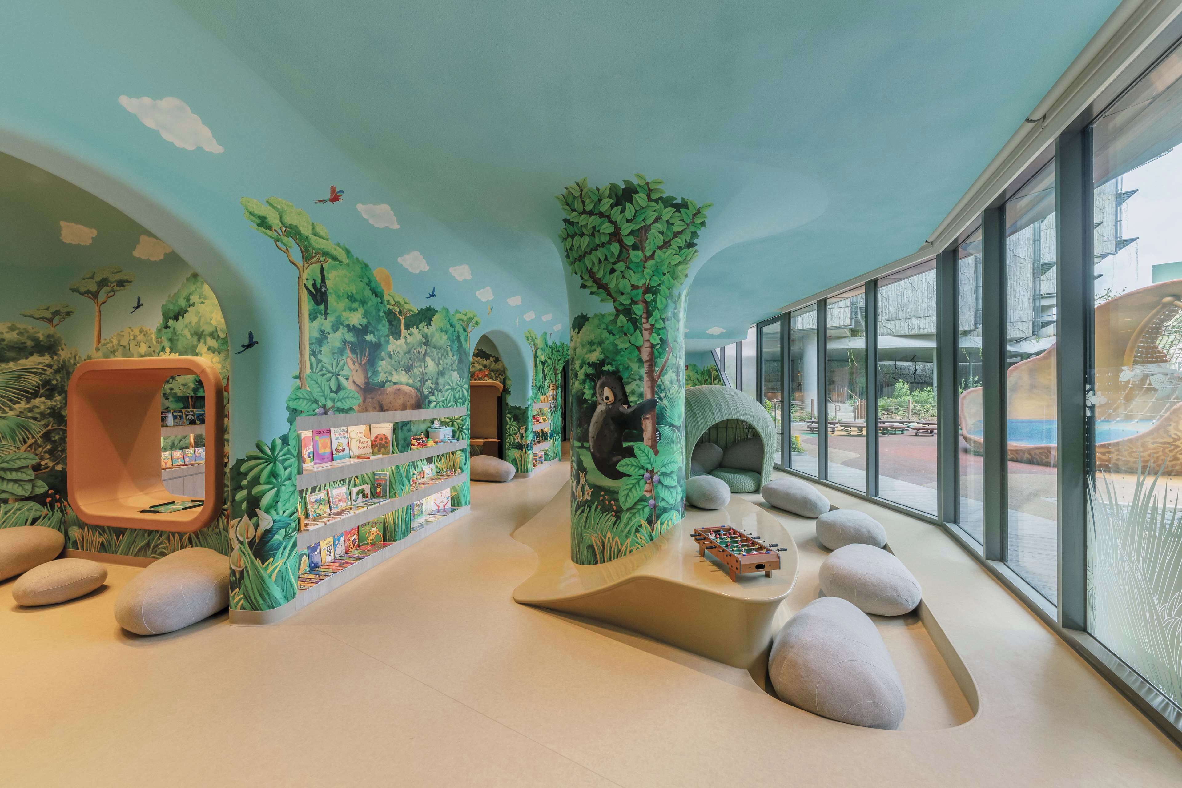 Espaço de recreação infantil com tema de selva, incluindo estante, áreas de estar e uma grande janela.