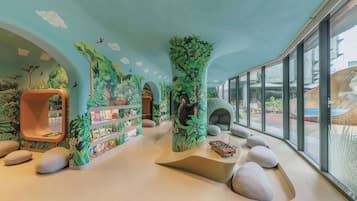 Espaço de recreação infantil com tema de selva, incluindo estante, áreas de estar e uma grande janela.