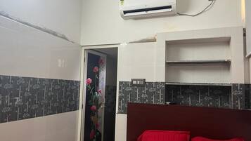 Quarto com cama vermelha, paredes brancas, ar condicionado branco e porta que dá para um banheiro com um balde rosa.