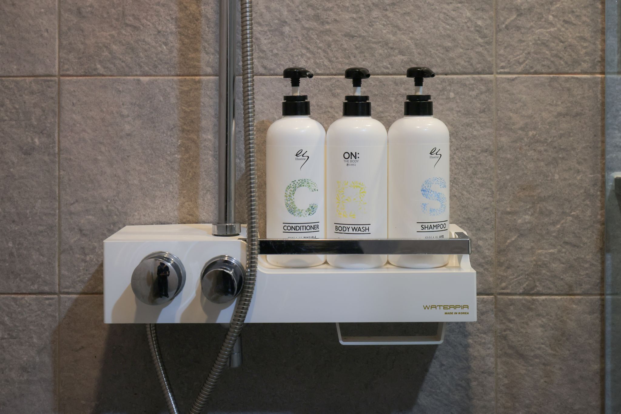 Uma área de chuveiro com três frascos de produtos à base de água, identificados como condicionador, hidratante corporal e shampoo.