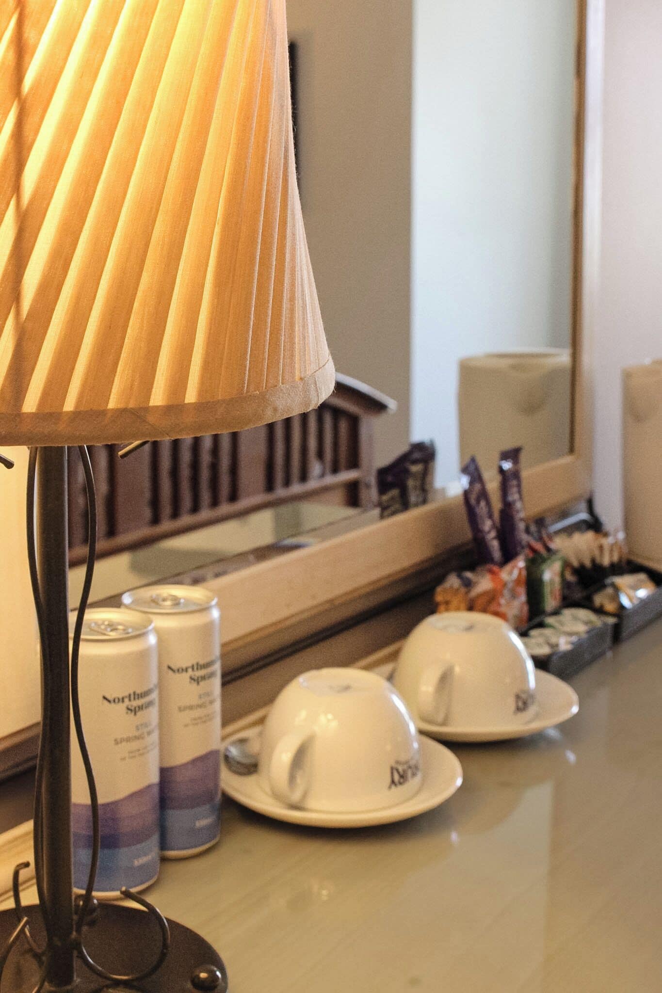 Quarto de hotel com abajur, espelho e minibar com petiscos e bebidas.
