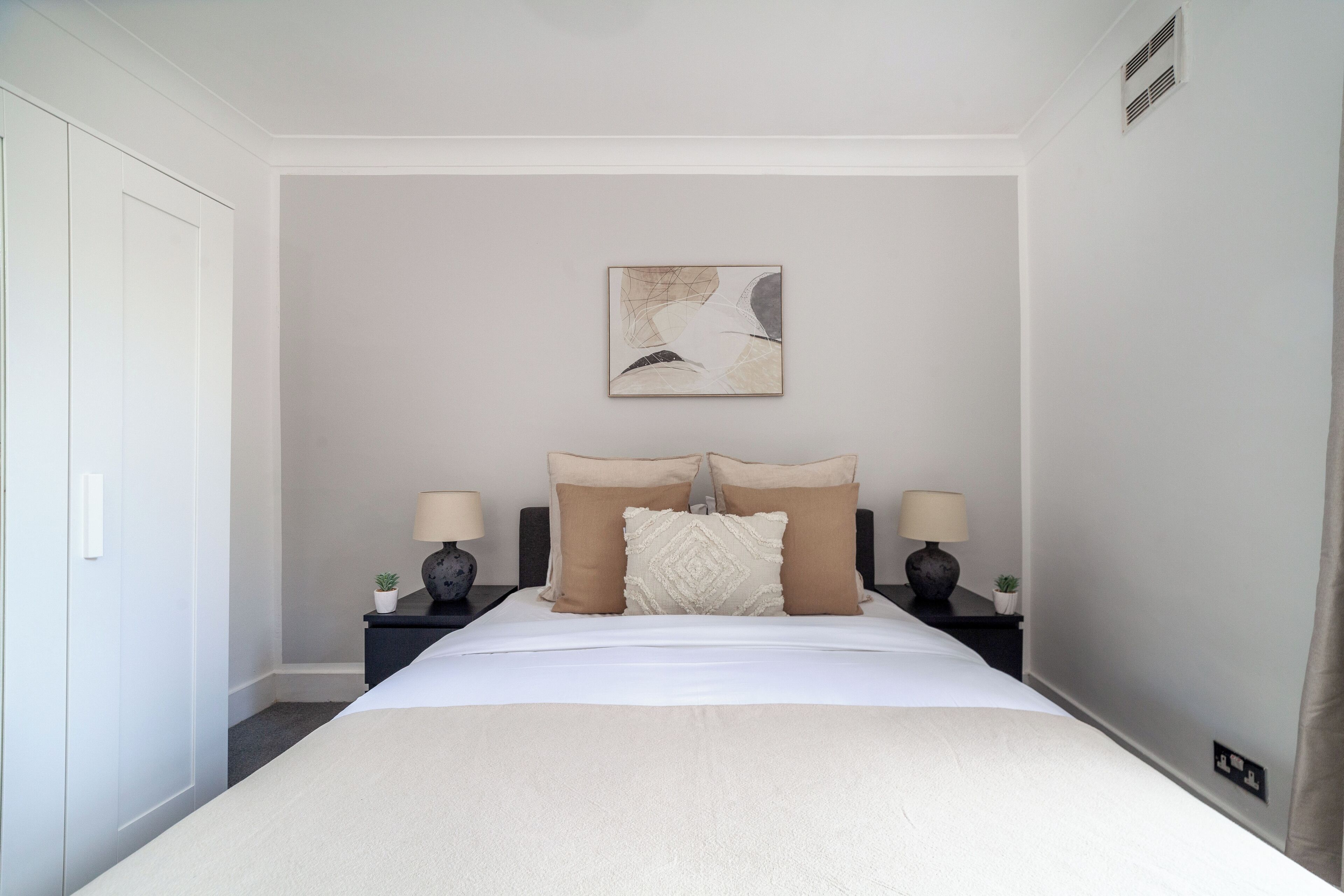 Quarto bem arrumado com uma cama grande, duas mesas de cabeceira com abajures, uma obra de arte na parede e um guarda-roupa branco.