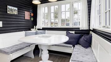 Quarto com mesa redonda branca e bancos, janela com cortinas brancas e parede de madeira preta.
