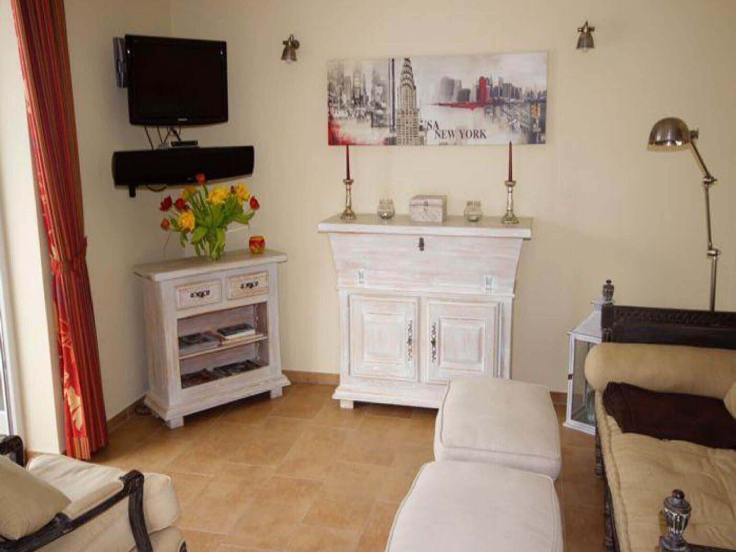 Uma sala de estar com um sofá, uma TV fixada na parede, um armário branco com um quadro e um abajur.