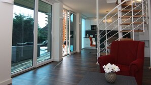 Uma sala de estar moderna com uma poltrona vermelha, uma mesa de centro com um vaso, e uma escadaria que leva a um andar superior.