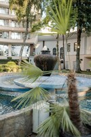 Uma fonte com bico, rodeada por uma piscina azulejada e palmeiras, com um edifício ao fundo.
