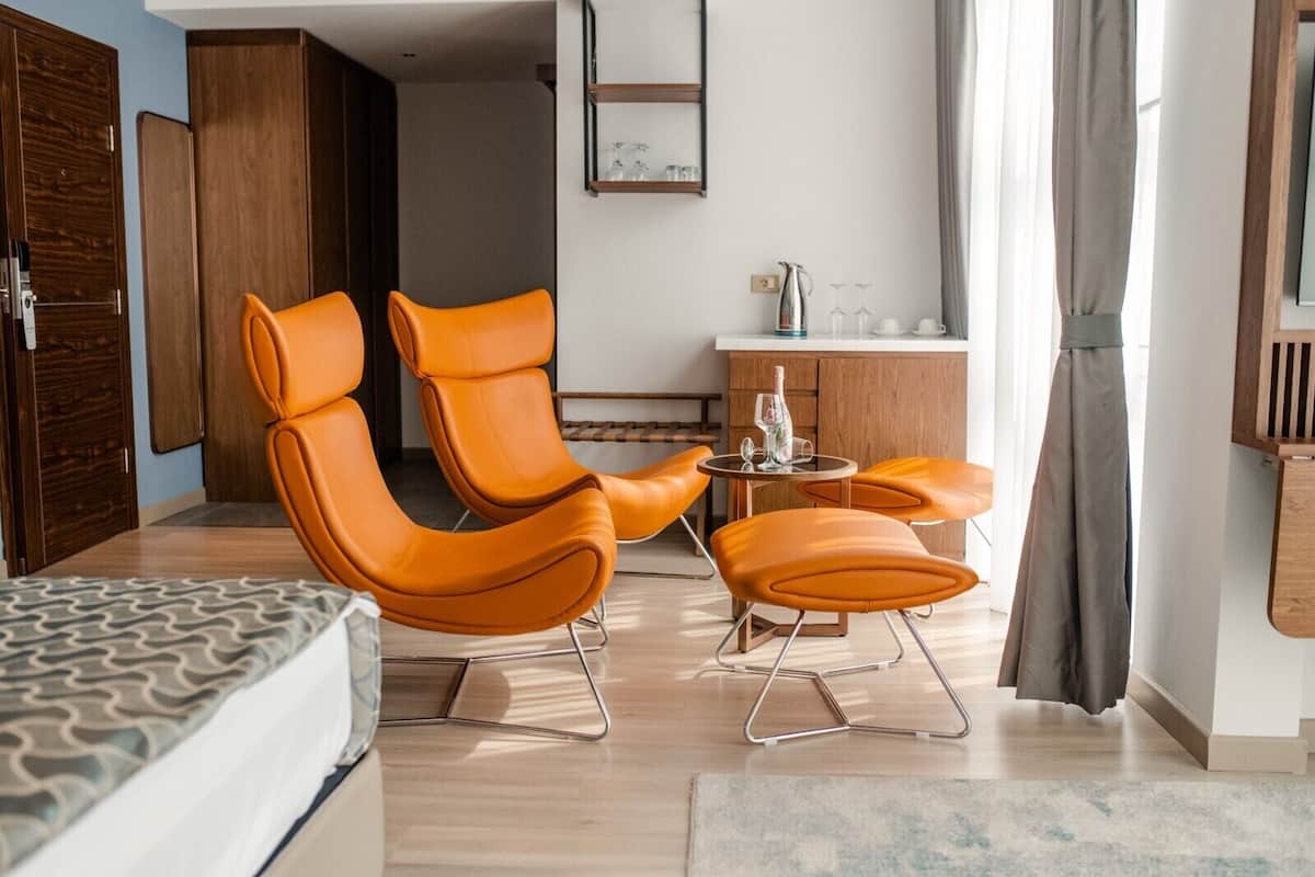Quarto de hotel moderno com uma cama, duas poltronas de couro laranja, uma mesinha redonda, um armário de madeira e um espelho.