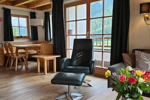 Quarto com mesa de jantar de madeira, uma cadeira de couro preto, um sofá e uma janela grande com cortinas.