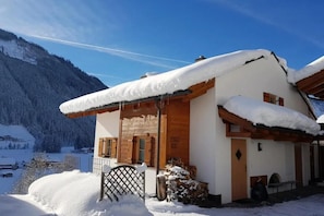 Um chalé coberto de neve, com fachada de madeira, varanda e montanhas ao fundo.