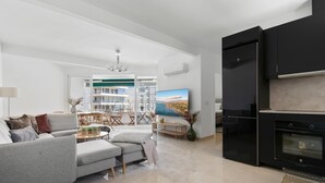 Uma sala de estar moderna com um sofá cinzento, uma mesa de centro e uma televisão. Há uma área de cozinha com um frigorífico e forno pretos.