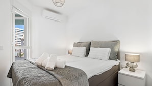 Quarto com cama, almofadas, mesa de cabeceira com abajur e vista para prédios por uma porta aberta.