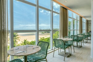 Restaurante com grandes janelas oferecendo vista para uma praia. Mesas e cadeiras dispostas para refeições.