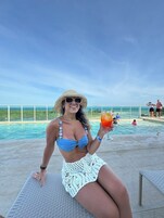 Uma pessoa de biquíni azul e saia de crochê branca sentada à beira da piscina, com uma bebida, tendo uma vista clara do oceano e uma cerca ao fundo.