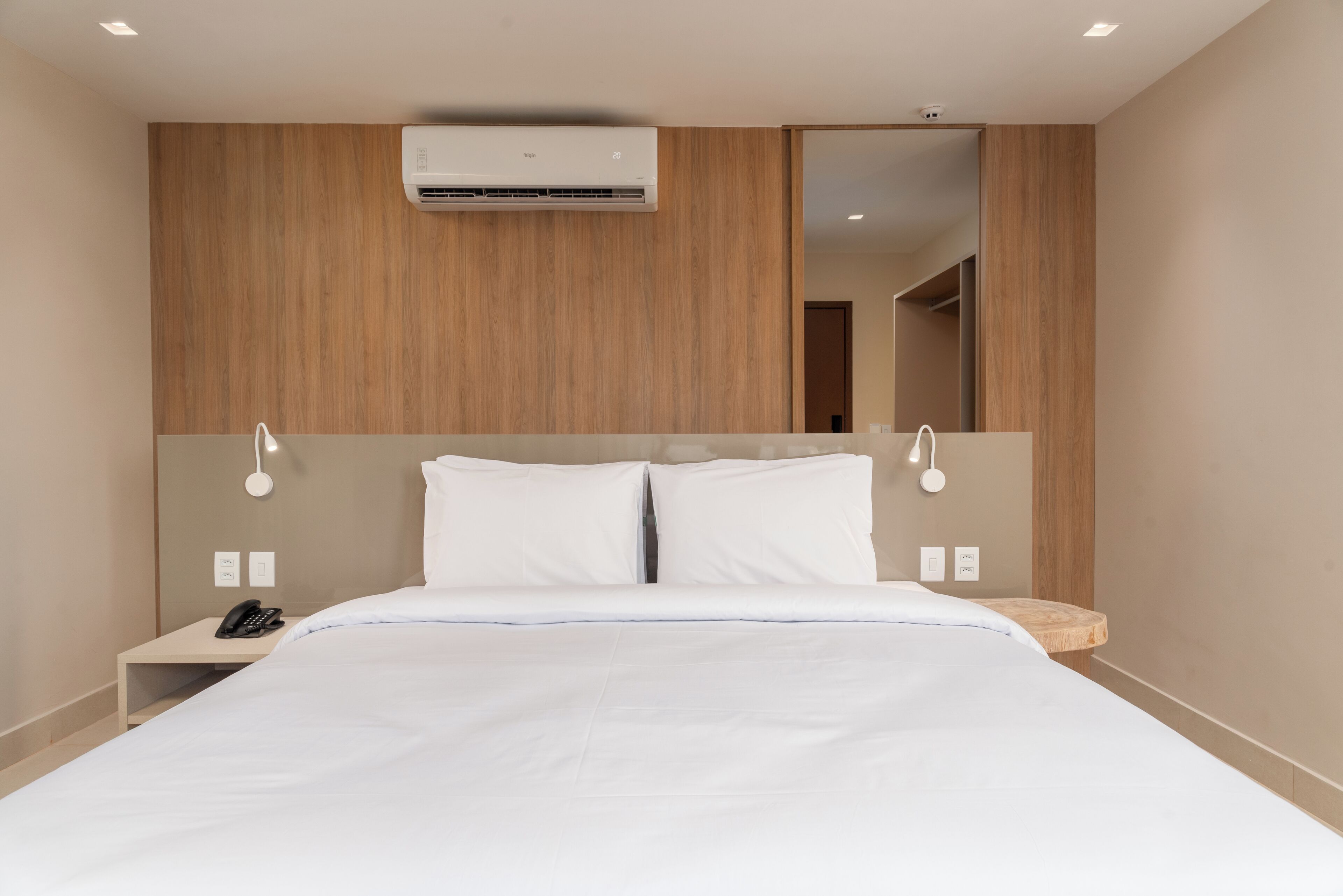 Quarto de hotel moderno com uma cama grande, painéis de madeira, ar condicionado embutido, abajures, um telefone na mesa de cabeceira e um espelho sobre a cama.