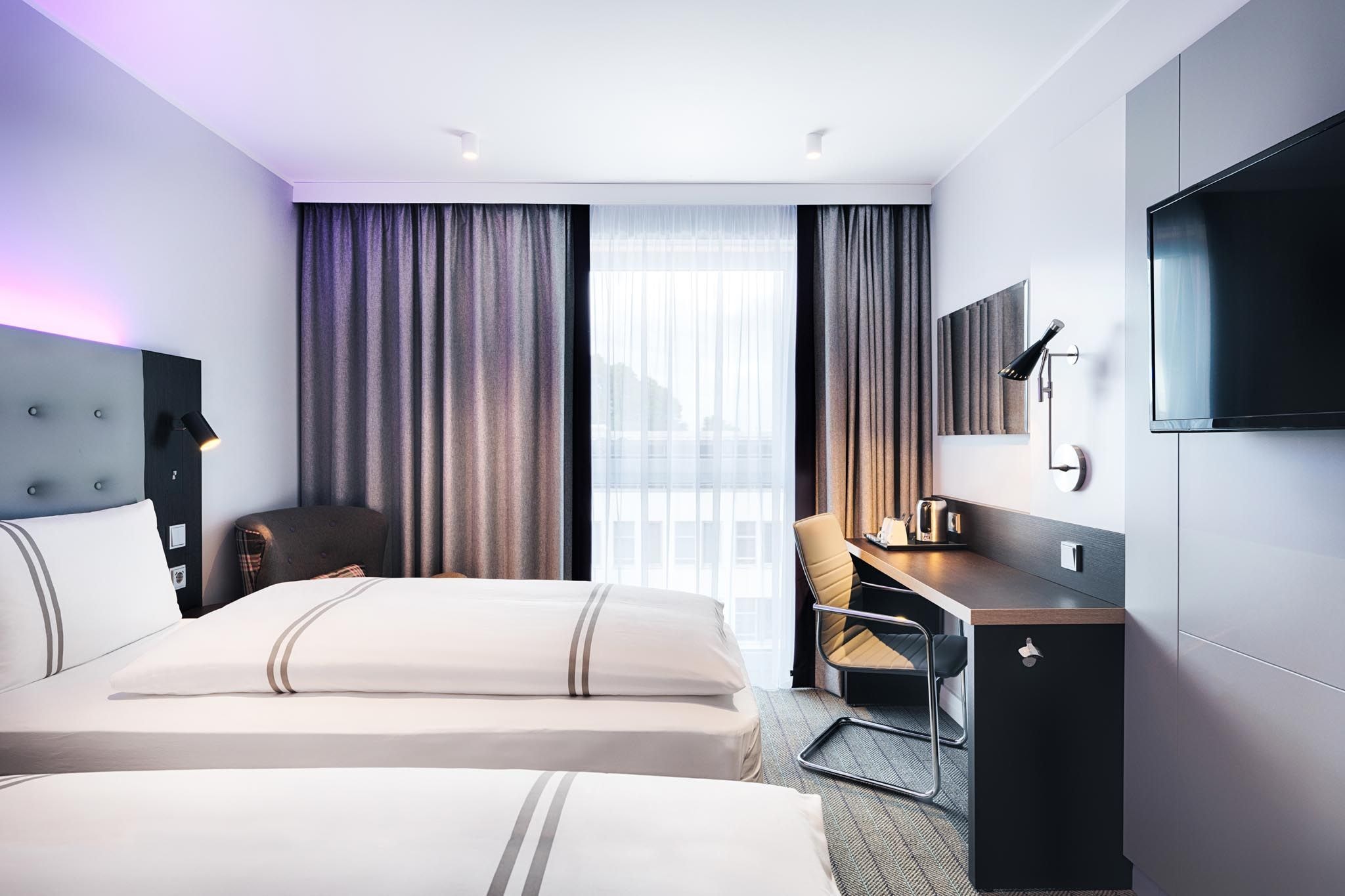 Quarto de hotel moderno com duas camas, uma escrivaninha, uma cadeira, televisão de tela plana e uma janela grande com cortinas.