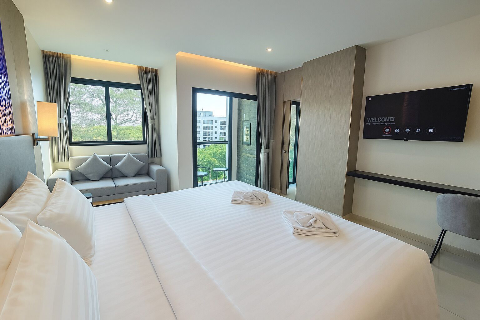 Quarto de hotel moderno com uma cama grande, um sofá, uma televisão e uma varanda com vista para prédios e árvores.