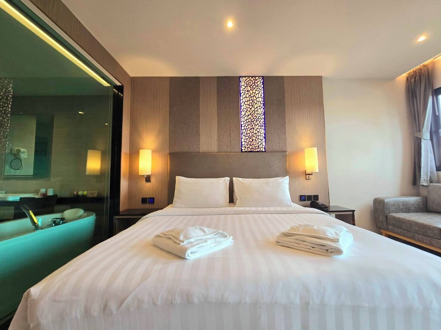 Quarto de hotel com cama grande, duas luminárias de cabeceira, um painel decorativo, banheiro com espelho e uma área de estar com uma poltrona.