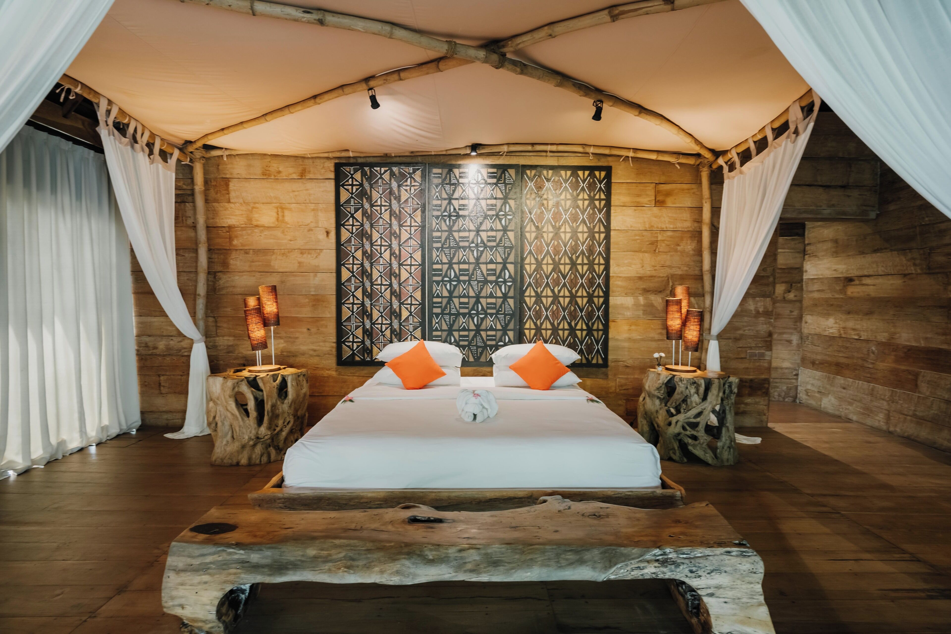 Uma cama grande com lençóis brancos e almofadas laranja, com duas mesas de cabeceira com luminárias, em um quarto com paredes de madeira e um dossel acima.