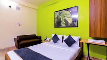Quarto de hotel com cama, sofá de couro marrom, parede verde e quadro com folhagens.