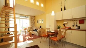 Uma sala de estar iluminada com sofá, mesa de jantar e cadeiras, uma cozinha com armários e bancada, e uma escada de madeira.
