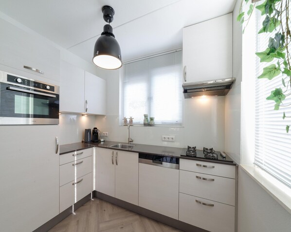 Uma cozinha moderna com armários brancos, eletrodomésticos de aço inoxidável, uma luminária pendente preta e uma janela com persianas.
