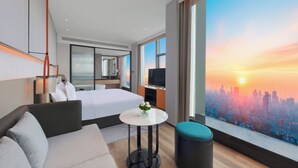 Quarto de hotel moderno com cama, sofá, mesa redonda e uma grande janela com vista para a cidade.