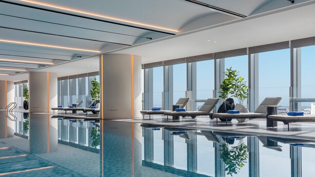 Uma piscina interna moderna com paredes de vidro transparente, cadeiras de descanso e vista para a cidade.