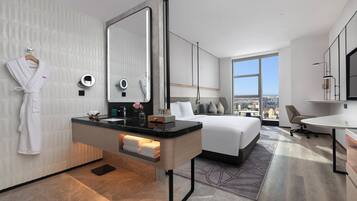 Quarto de hotel moderno com uma cama grande, uma toucador com espelho, um roupão pendurado na parede e vista para a cidade pela janela.