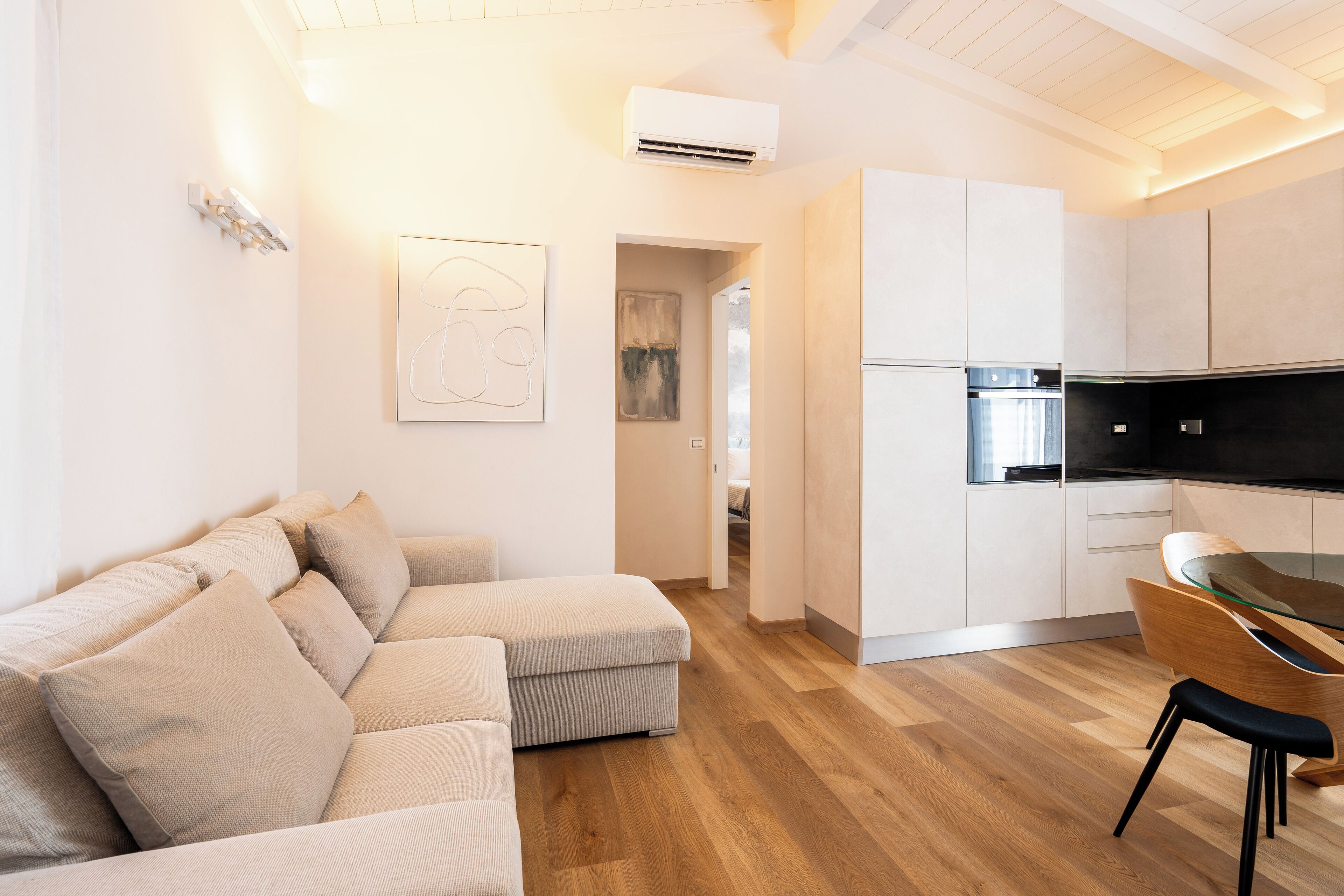 Uma sala moderna com sofá, área de jantar com mesa de vidro e cozinha com armários brancos e bancada preta.