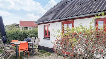 Uma casa branca com porta vermelha e janelas, telhado de telhas e um pequeno jardim com uma mesa e cadeiras.