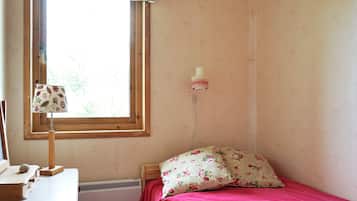 Quarto com uma cama de solteiro, mesa de cabeceira com abajur, radiador e janela com cortina florida.