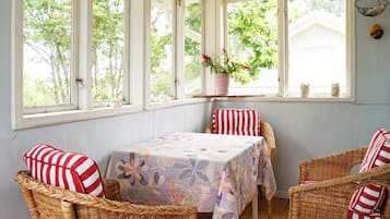 Um quarto com uma mesa coberta por uma toalha florida, cadeiras de vime com almofadas listradas, e uma janela com vista para árvores.