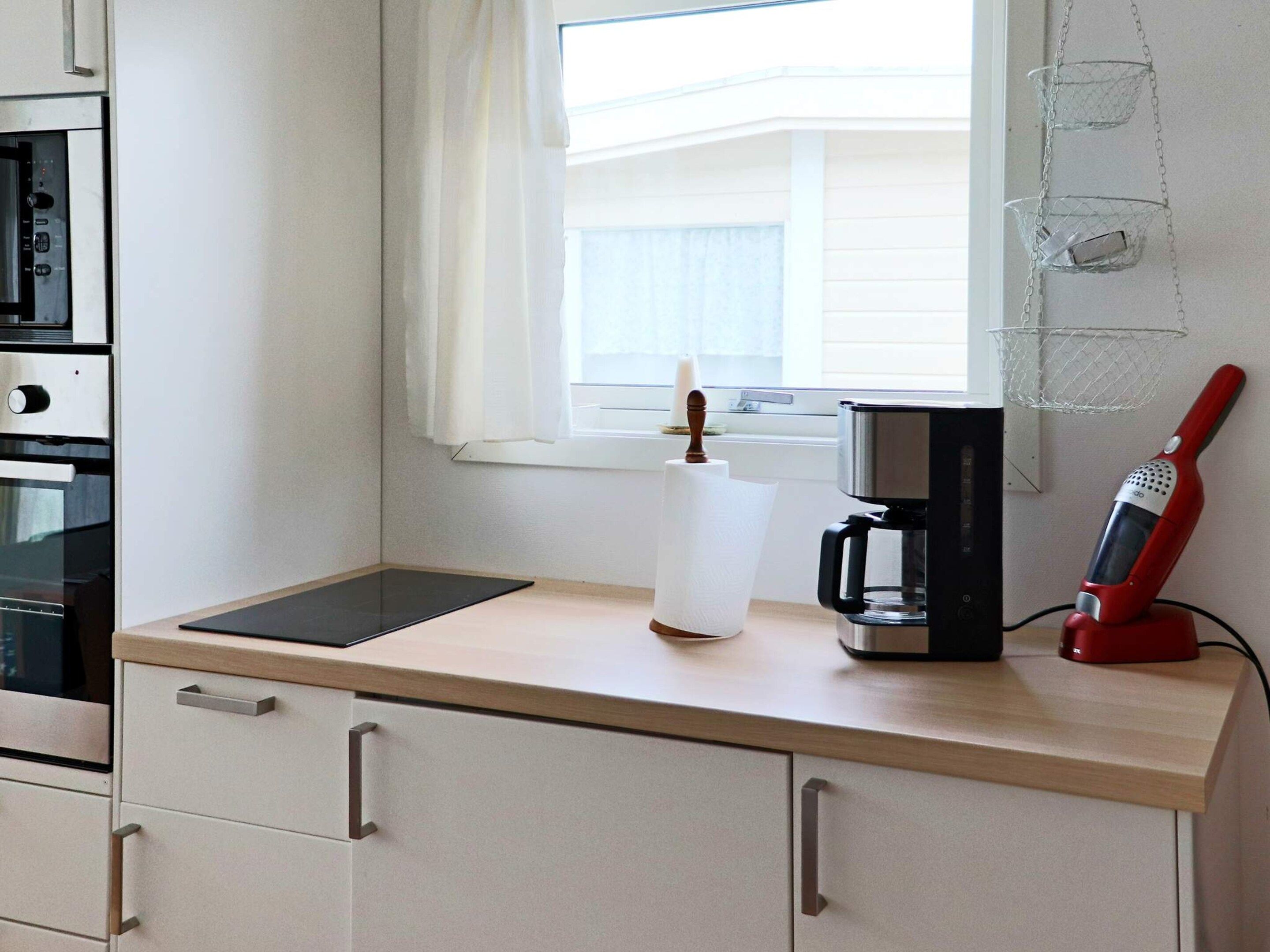 Cozinha com bancada, cafeteira, chaleira elétrica vermelha e janela com persianas.