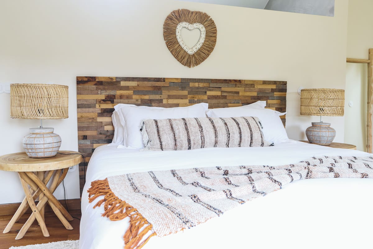 Quarto com cabeceira de madeira, duas mesas de cabeceira com abajures de fibra, um ornamento de parede em formato de coração e um cobertor estampado.