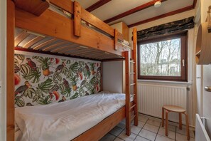 Uma cama beliche com escada de madeira, um banquinho pequeno e uma janela com vista para árvores.