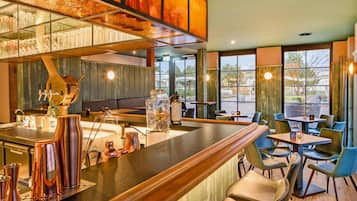 Uma área de bar moderna com balcão de madeira, copos de cobre e uma vitrine de vidro. Há áreas de estar com cadeiras azuis e vista para o exterior através de grandes janelas.