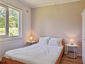 Um quarto com uma janela grande, uma cama com roupa de cama branca, duas mesas de cabeceira com abajures e um radiador sob a janela.