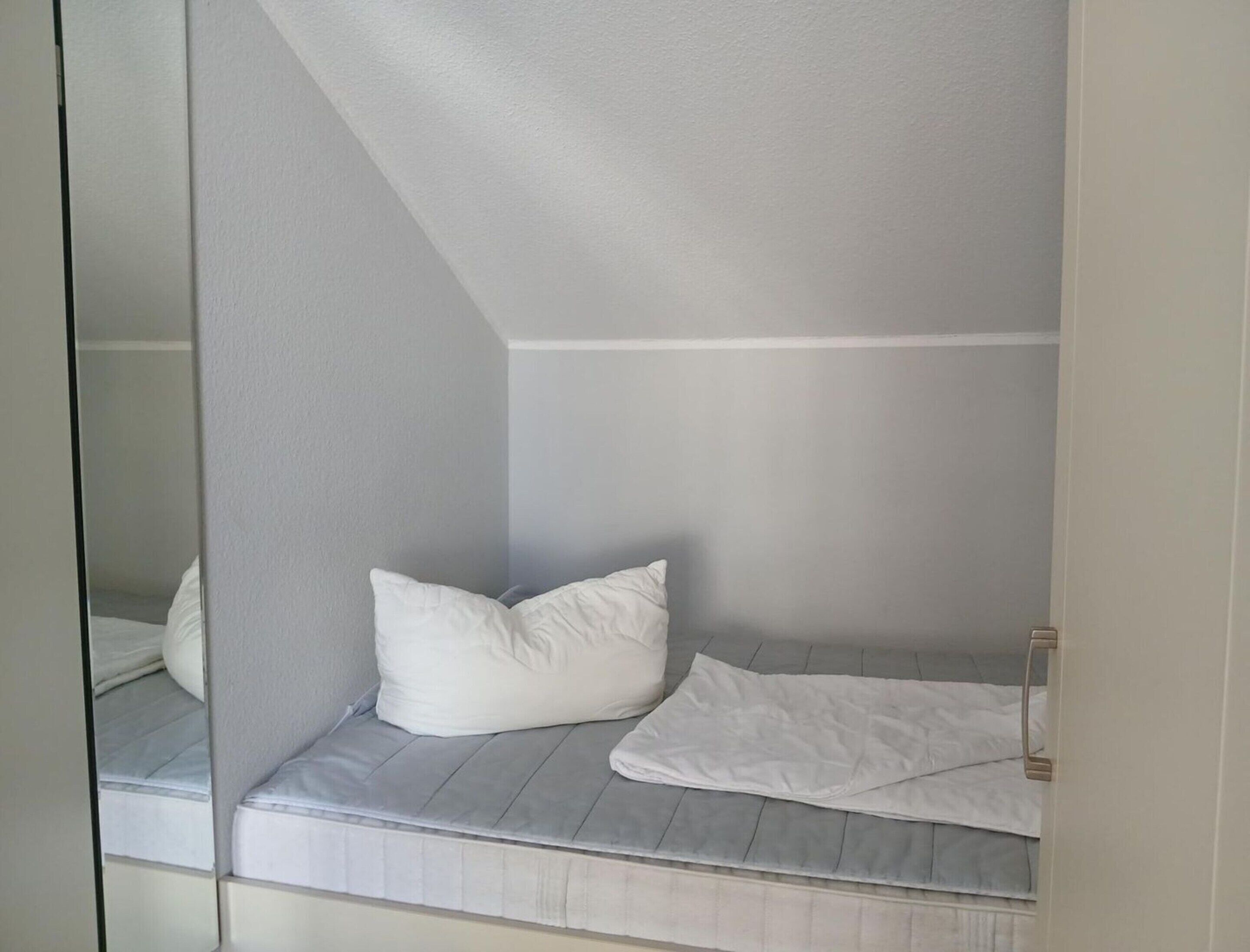Quarto com teto inclinado, cama com roupa de cama branca e janela com vista.