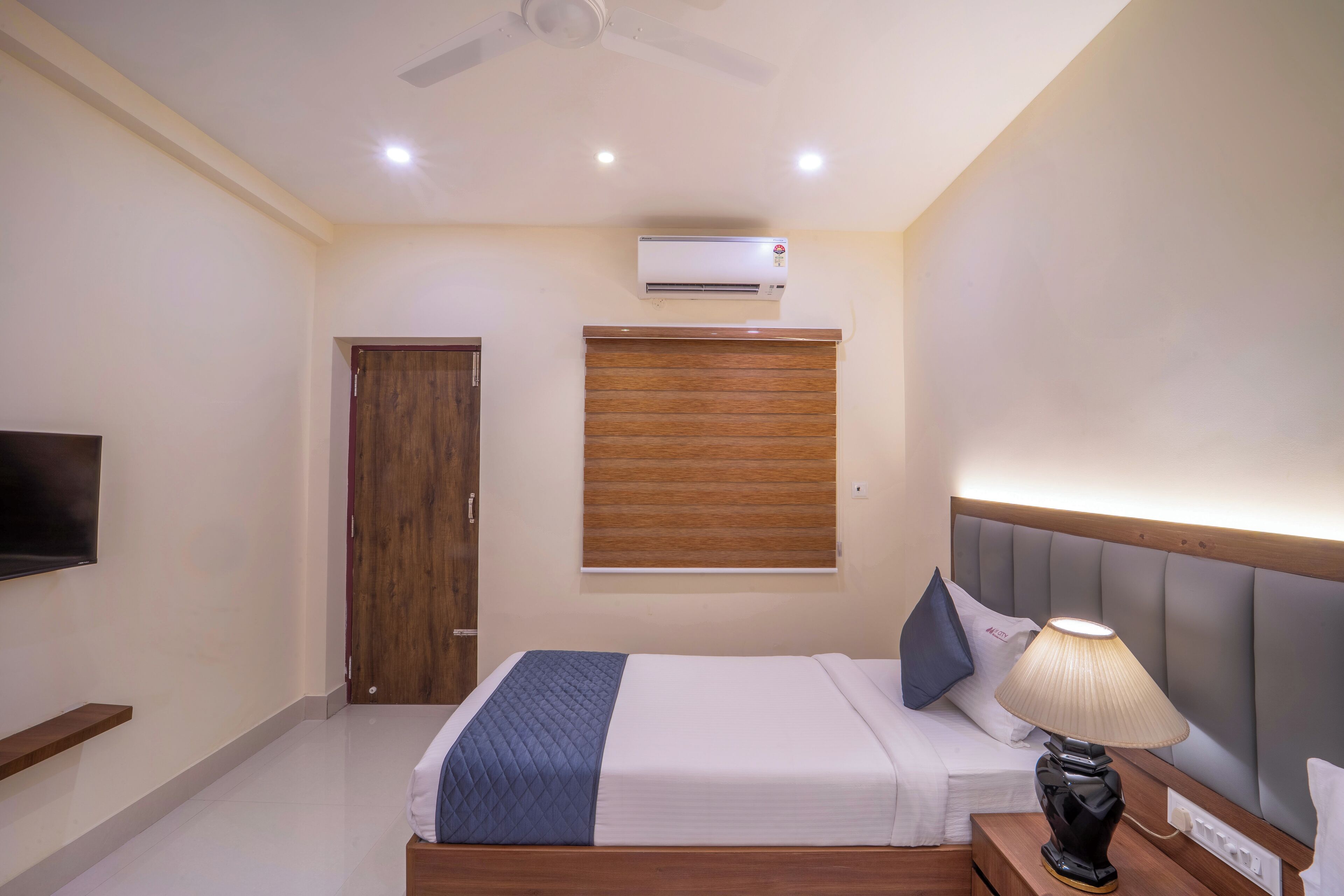 Quarto de hotel com cama, luminária, ar condicionado e televisão na parede.
