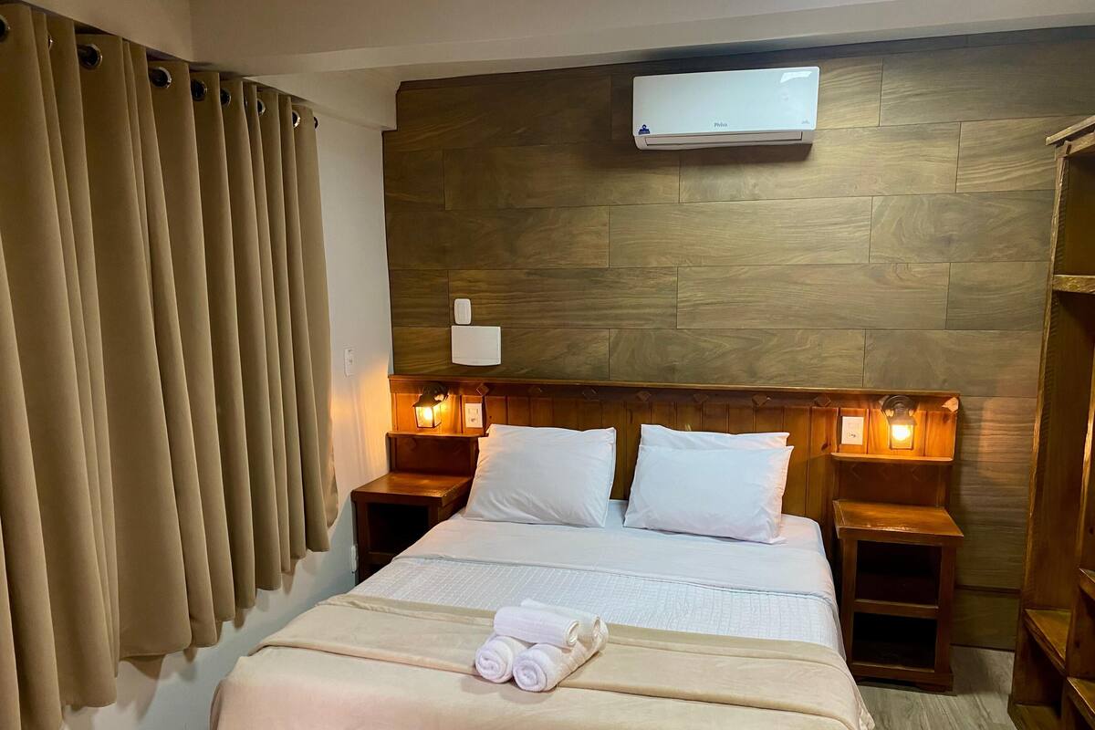Quarto de hotel com cama, duas mesinhas de cabeceira, ar condicionado fixado na parede e janela com cortinas.