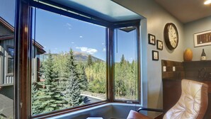 Quarto com uma janela grande que oferece vista para árvores e uma montanha. Há uma poltrona de couro e uma mesa pequena.