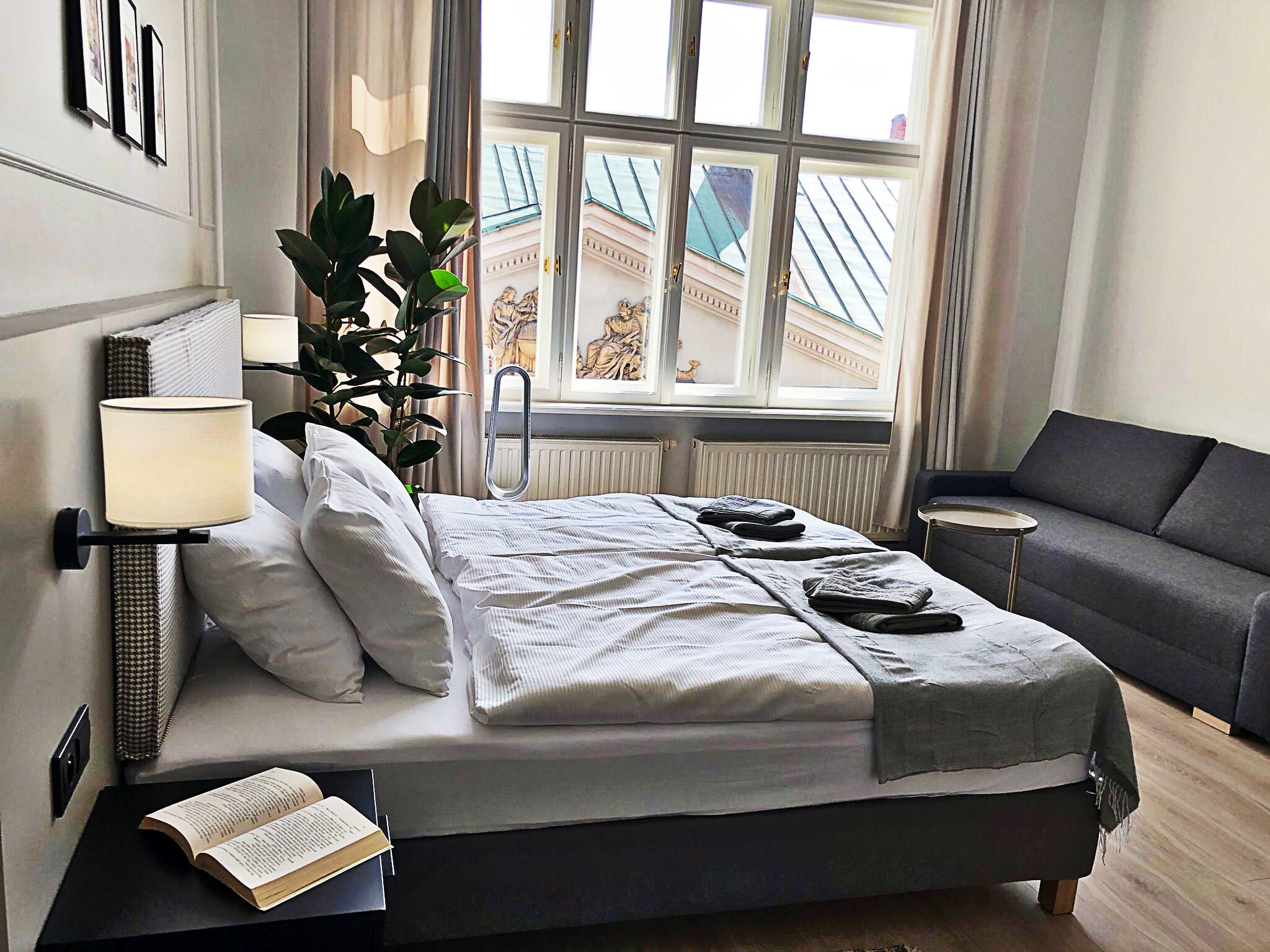 Quarto de hotel moderno com uma cama grande, uma mesa de cabeceira com um livro, uma mesinha redonda, um sofá cinzento e uma janela com vista para edifícios.